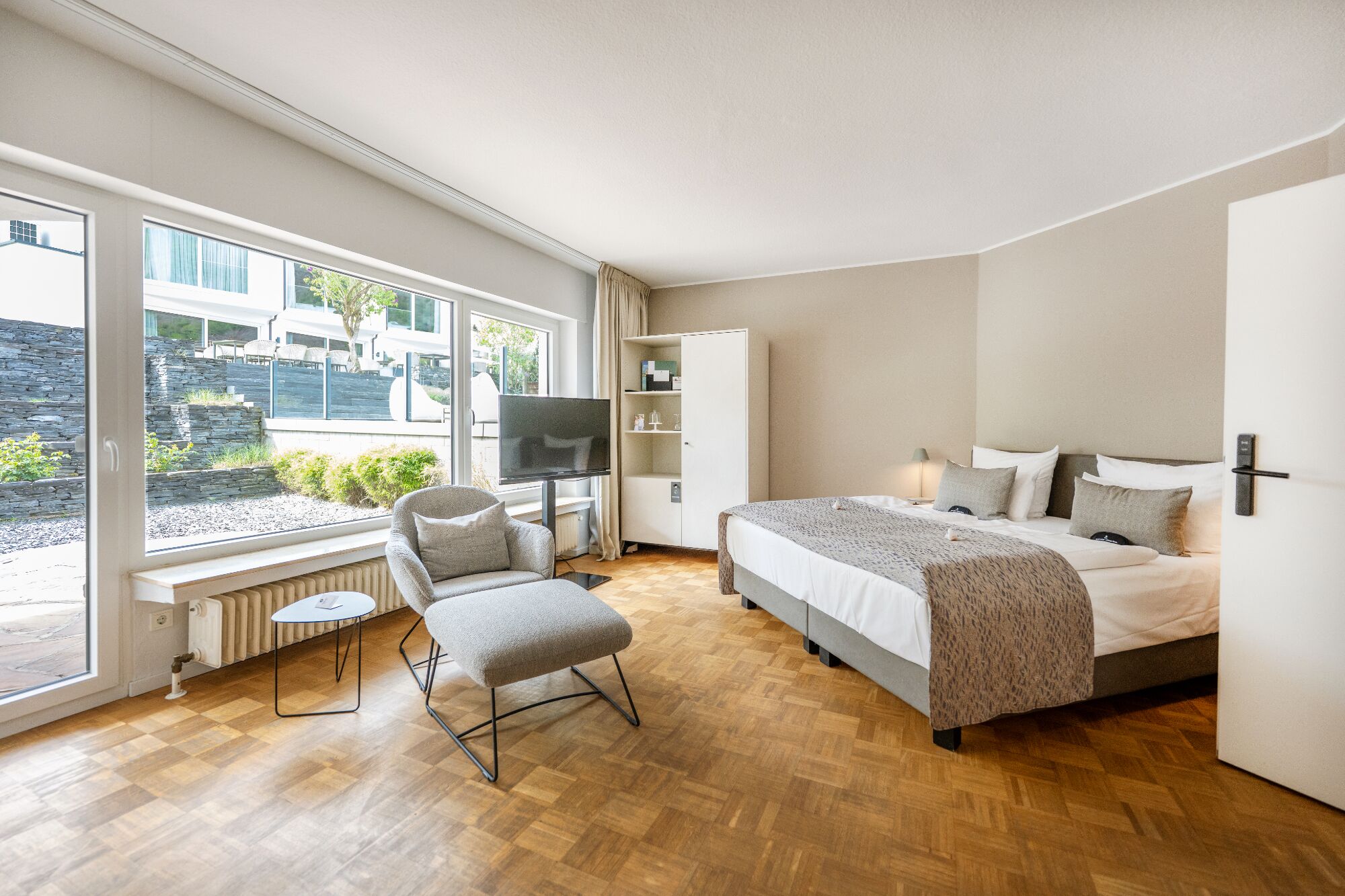 Quarto de hotel moderno com uma cama grande, televisão, área de estar com uma poltrona e um banquinho, e vista para o exterior.