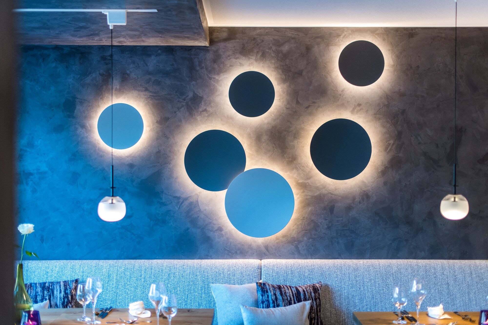 Um interior moderno com luminárias circulares na parede e uma área de jantar com parede texturizada em azul e mesa de madeira.