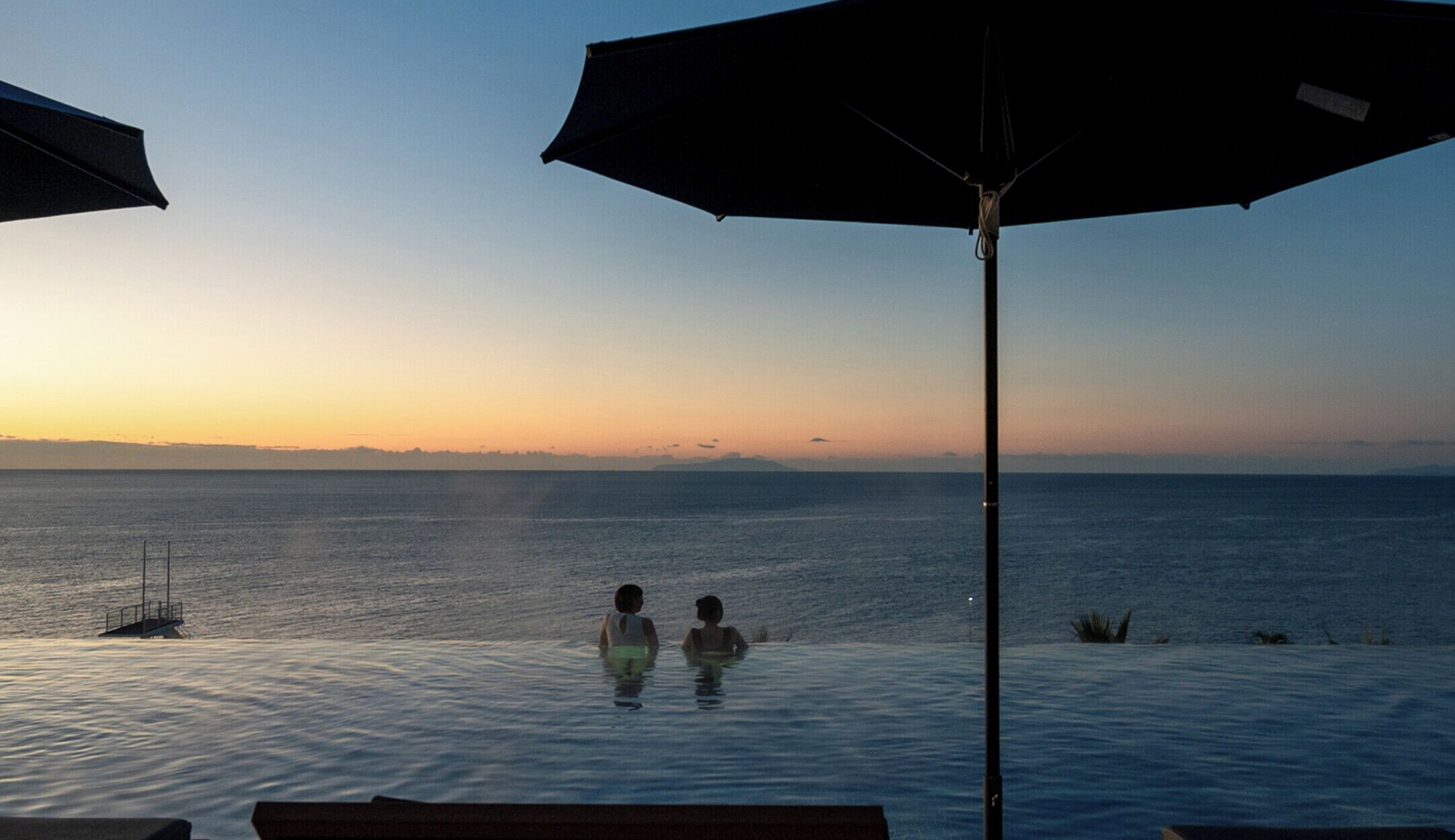 Piscina infinita com vista para o mar e montanhas ao longe durante o pôr do sol.