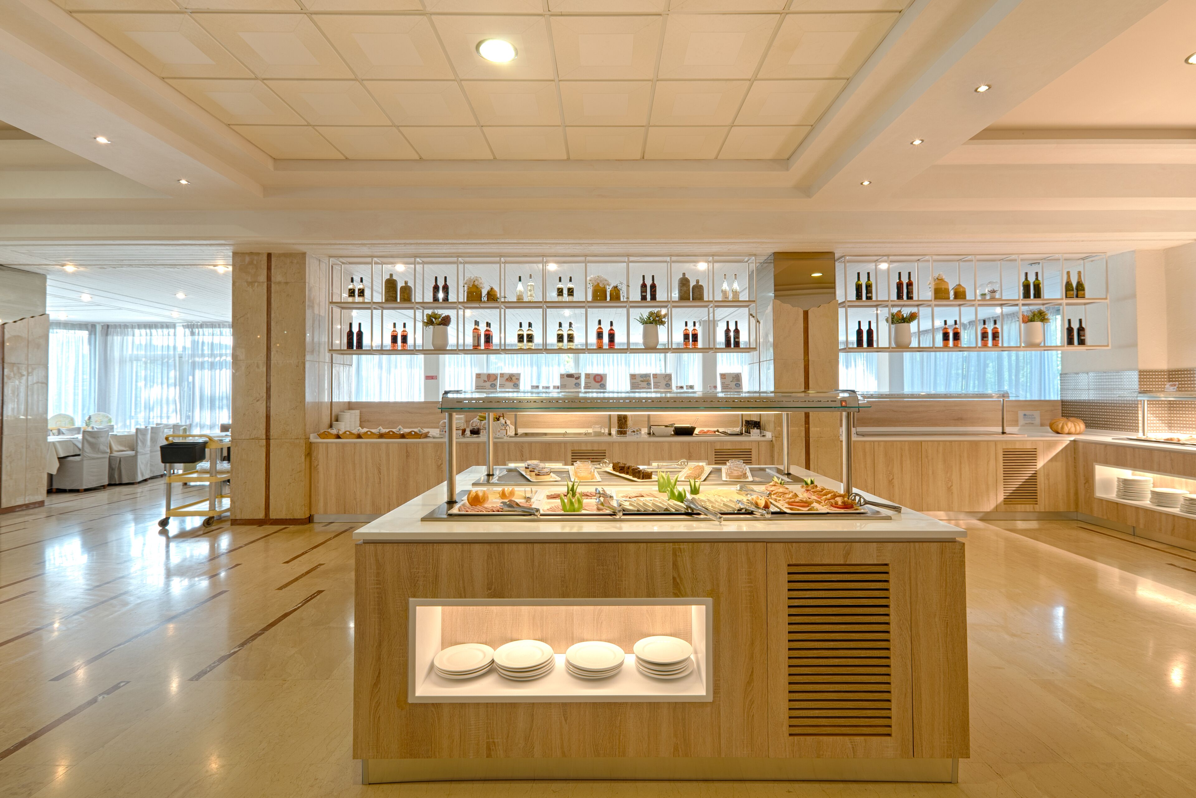 Uma área de refeições moderna em hotel, com serviço de buffet, painéis de madeira e diversas opções de comida e bebida.