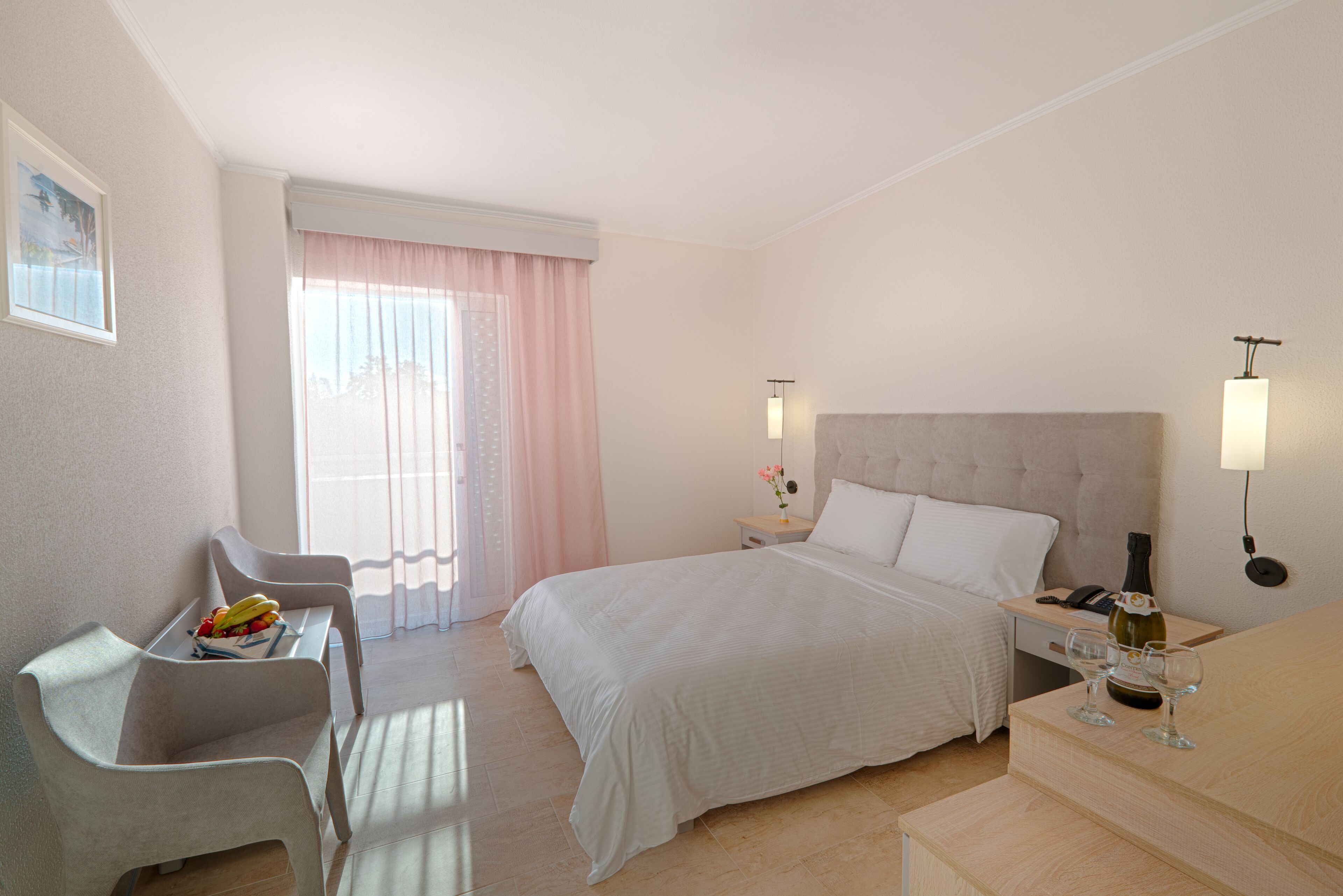 Quarto de hotel com cama, escrivaninha com cadeira, mesa pequena com frutas e janela com cortinas.