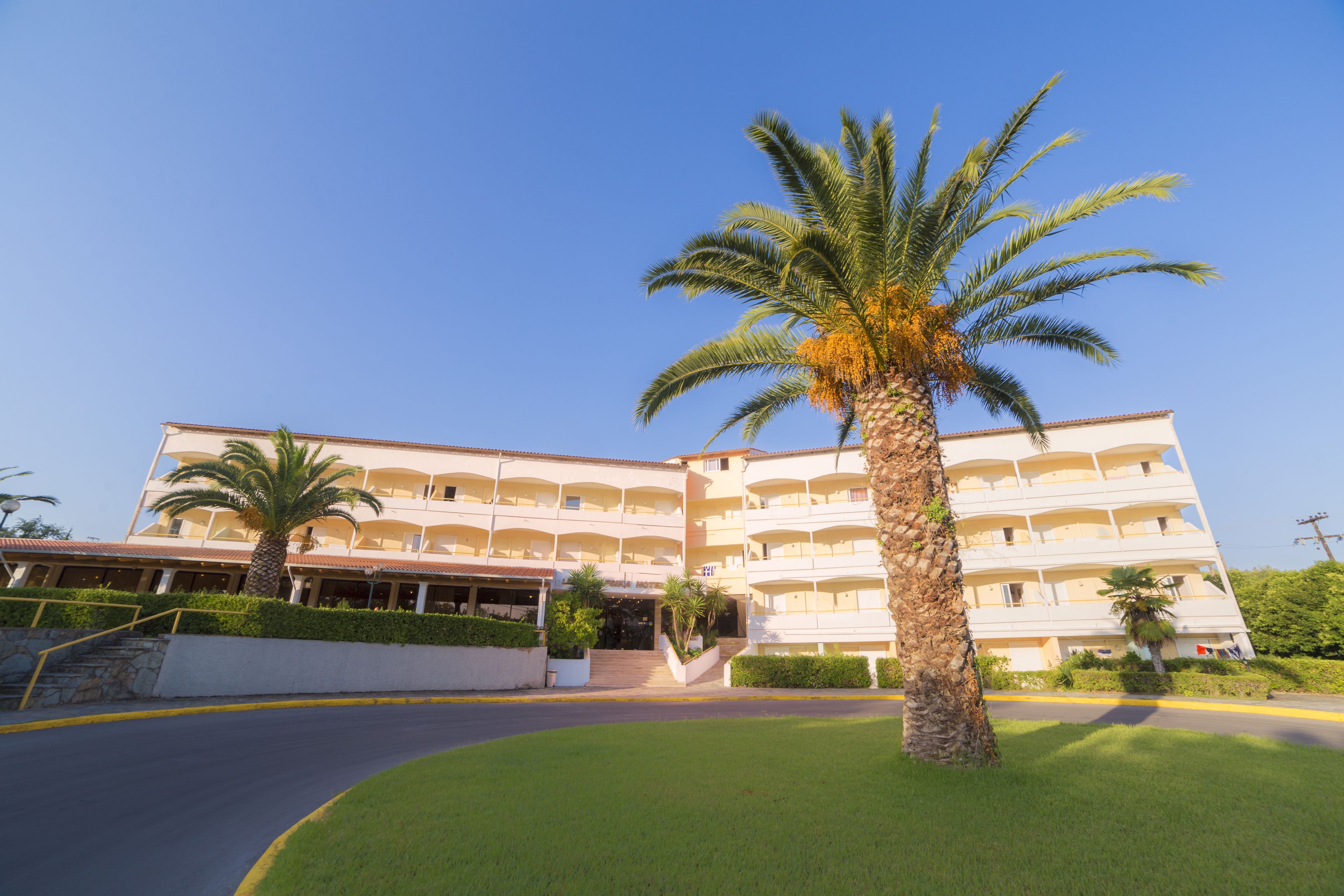 Hotel com vários andares, entrada central, palmeiras e céu azul claro.