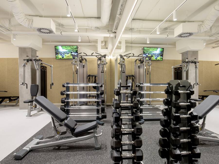 Uma academia com equipamentos de musculação, aparelhos de ginástica e uma tela de televisão na parede.
