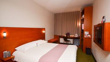 Quarto de hotel com uma cama grande, uma escrivaninha, uma cadeira, uma mesinha de centro, um cesto de lixo e uma televisão.
