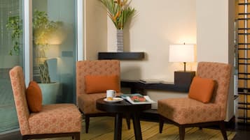 Quarto de hotel com duas poltronas estampadas, uma mesa pequena com uma xícara e um livro, um vaso com flores e um abajur.