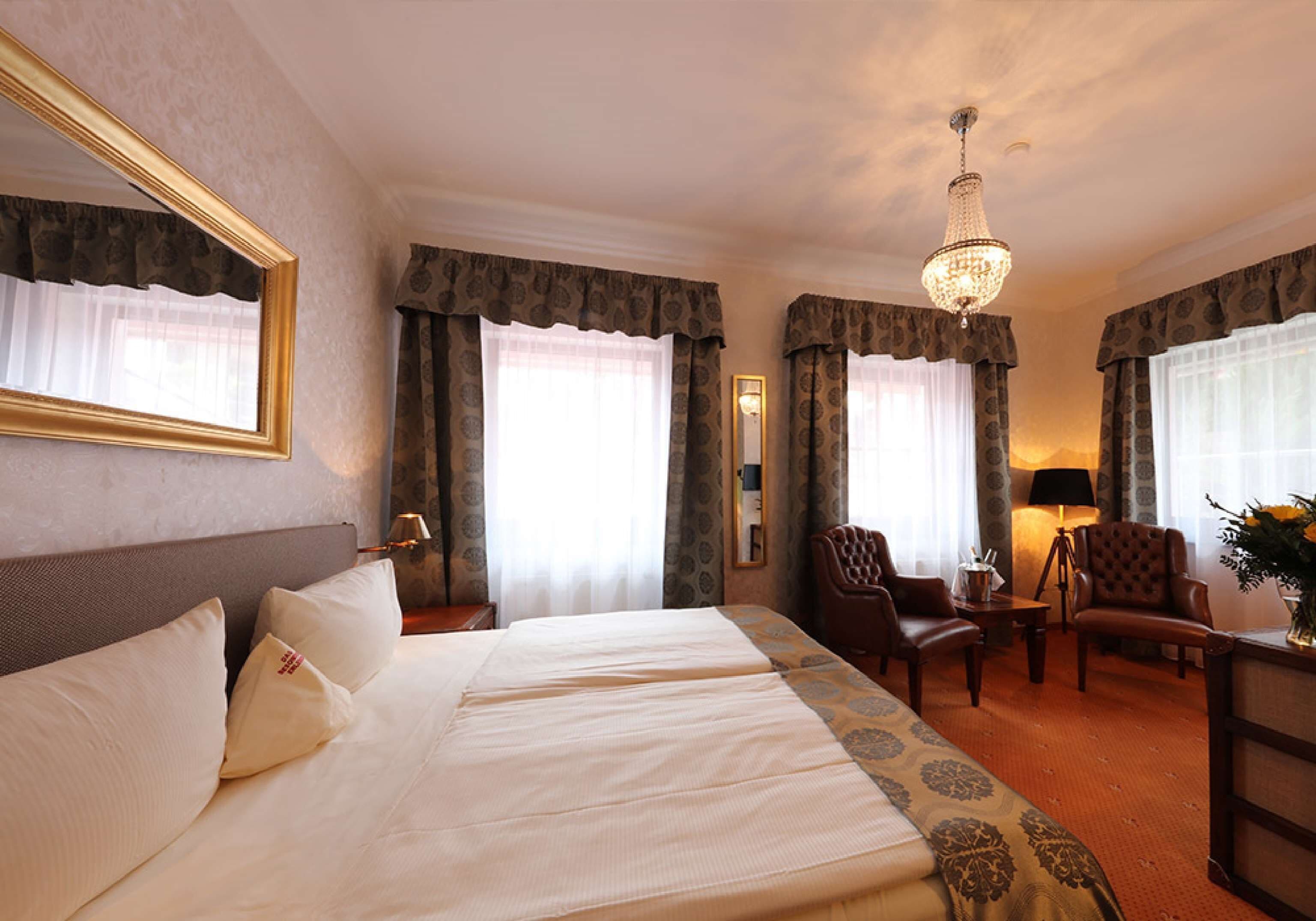 Quarto de hotel com cama grande, duas poltronas, escrivaninha e lustre.
