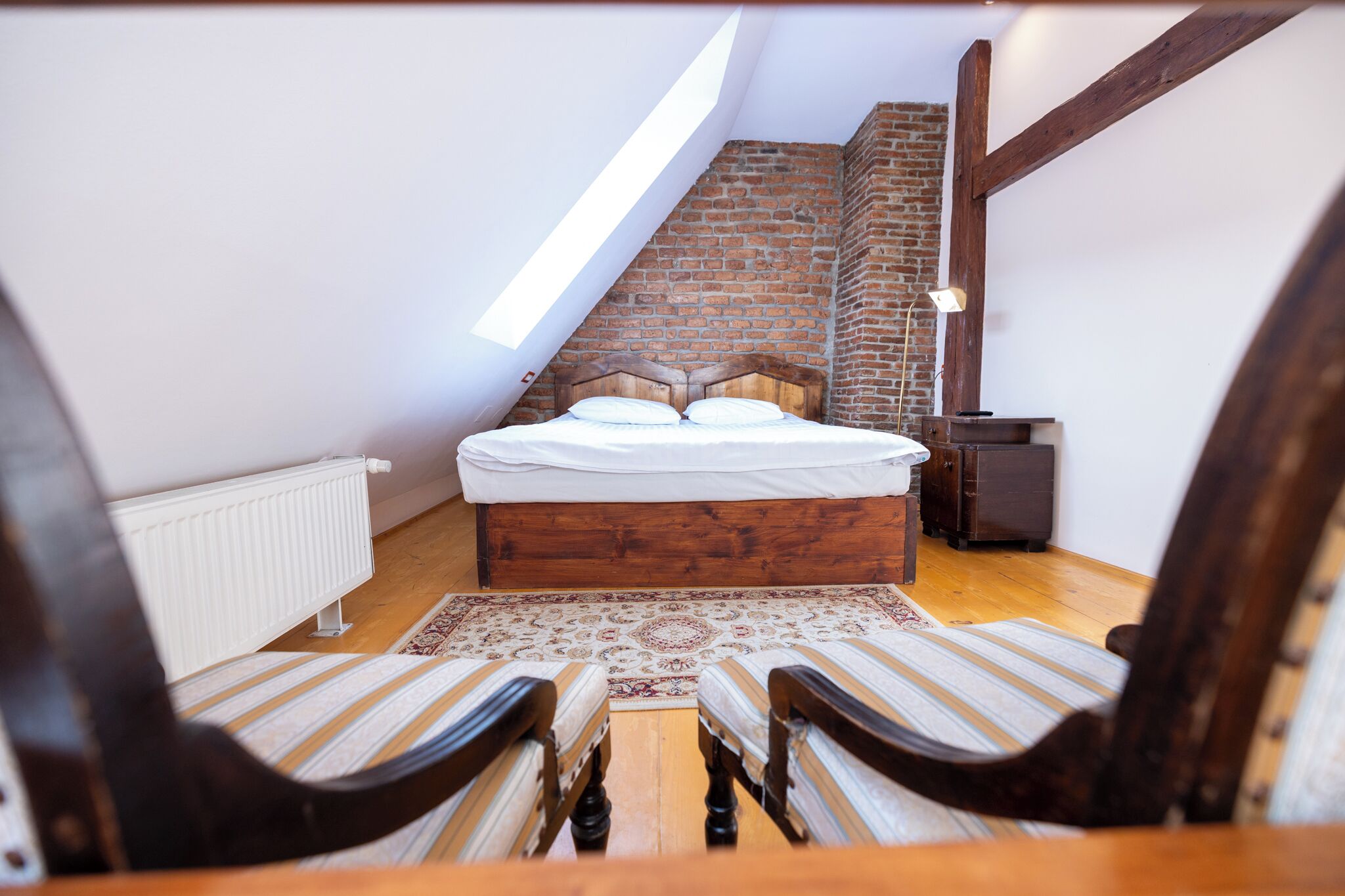 Quarto estilo loft com teto inclinado, parede de tijolos, cama de madeira, radiador branco, tapete estampado e duas poltronas estofadas.