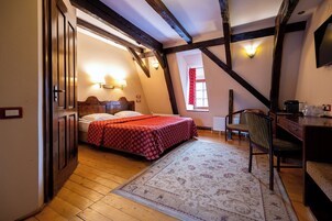 Um quarto com piso de madeira, uma cama com colcha vermelha estampada, uma escrivaninha com cadeira e uma janela com cortinas vermelhas.