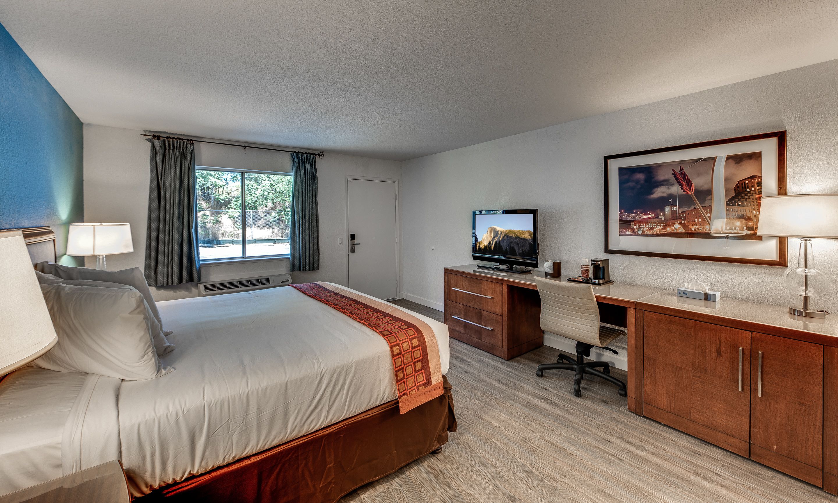 Quarto de hotel com cama, escrivaninha, cadeira, televisão e um quadro na parede.