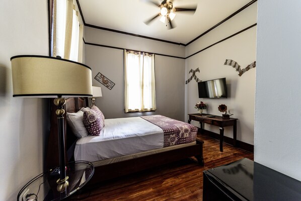 Quarto com cama, mesa de cabeceira, luminária, ventoinha no teto, janela com cortinas e televisão fixada na parede.