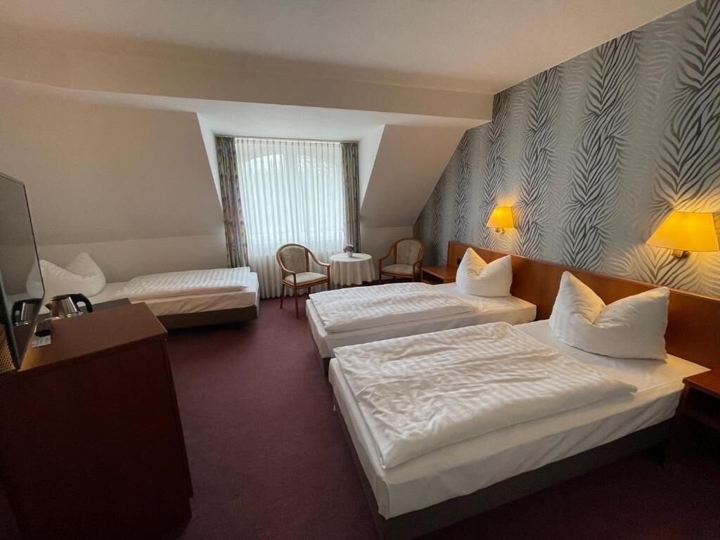 Quarto de hotel com duas camas, uma mesa, uma cadeira, uma mesinha e uma janela com cortinas.