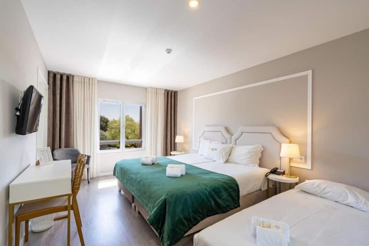 Quarto de hotel com duas camas, uma escrivaninha, uma cadeira, televisão e janela com cortinas.