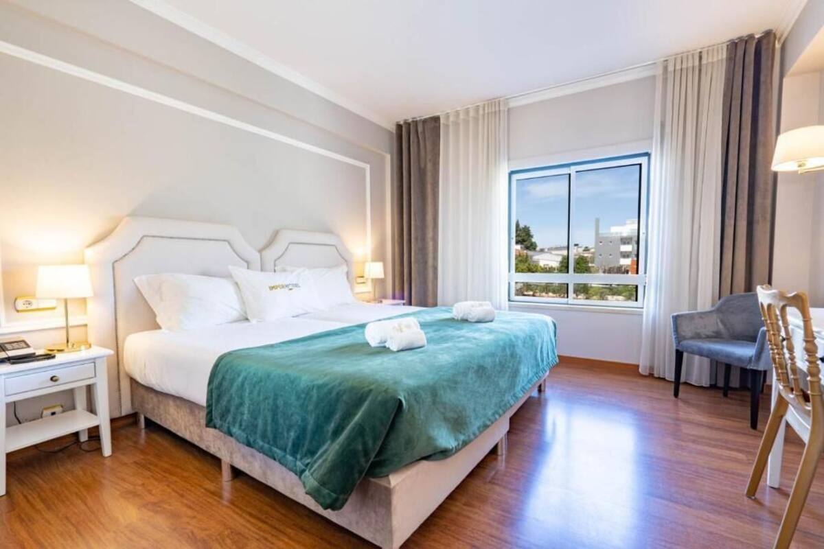 Quarto de hotel com uma cama grande, mesa de cabeceira, cadeira e janela com vista para a cidade.