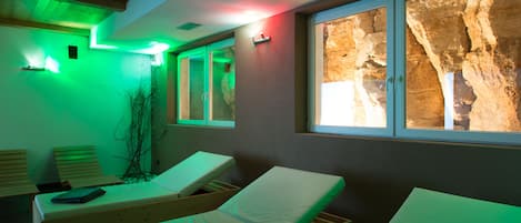 Sala de spa com espreguiçadeiras de madeira, uma janela ampla com vista para pedras e iluminação ambiente esverdeada.