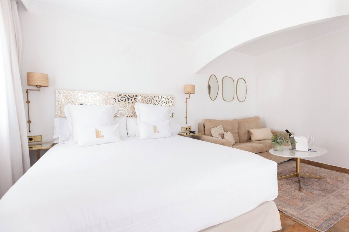 Quarto de hotel com uma cama grande, um sofá, uma mesinha com uma garrafa e copos, e dois espelhos fixados na parede.