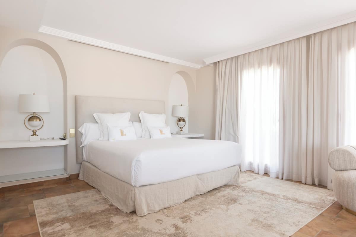 Uma cama grande com lençóis brancos, uma mesa de cabeceira com abajur e uma janela ampla com cortinas.