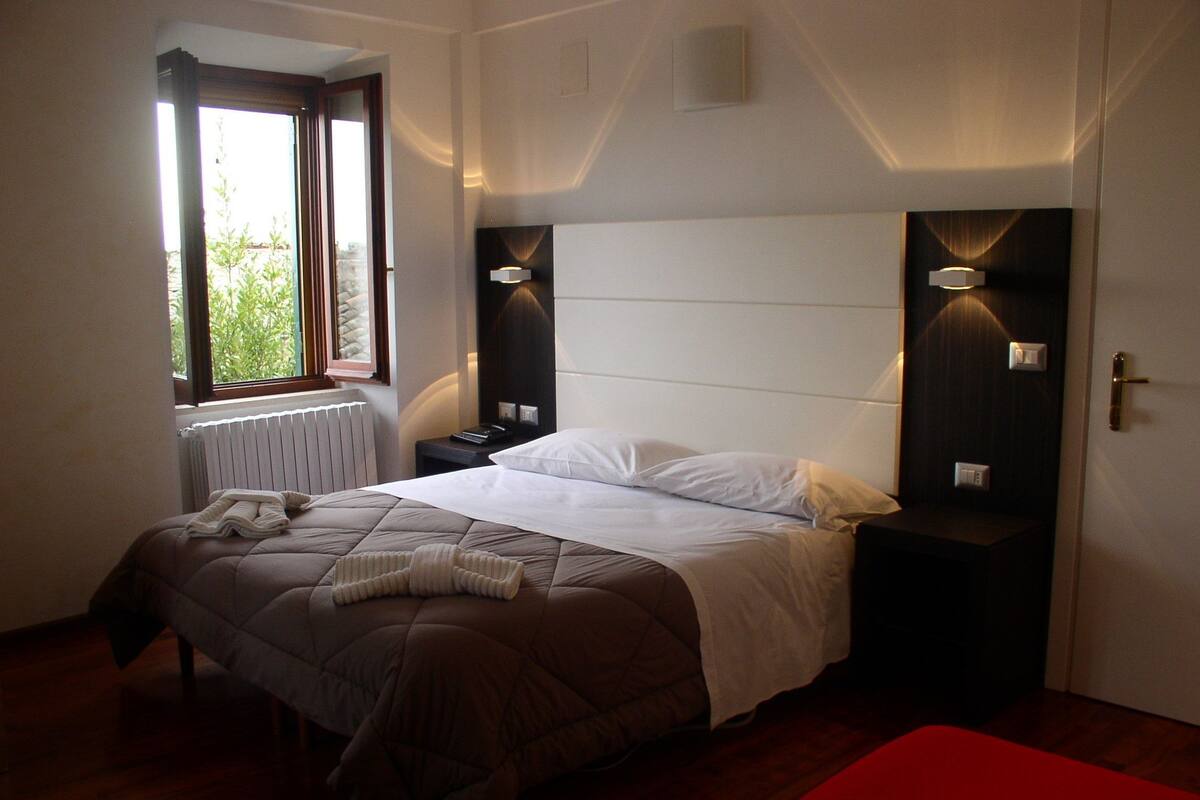 Quarto com cama, janela, radiador e duas mesas de cabeceira com abajures.