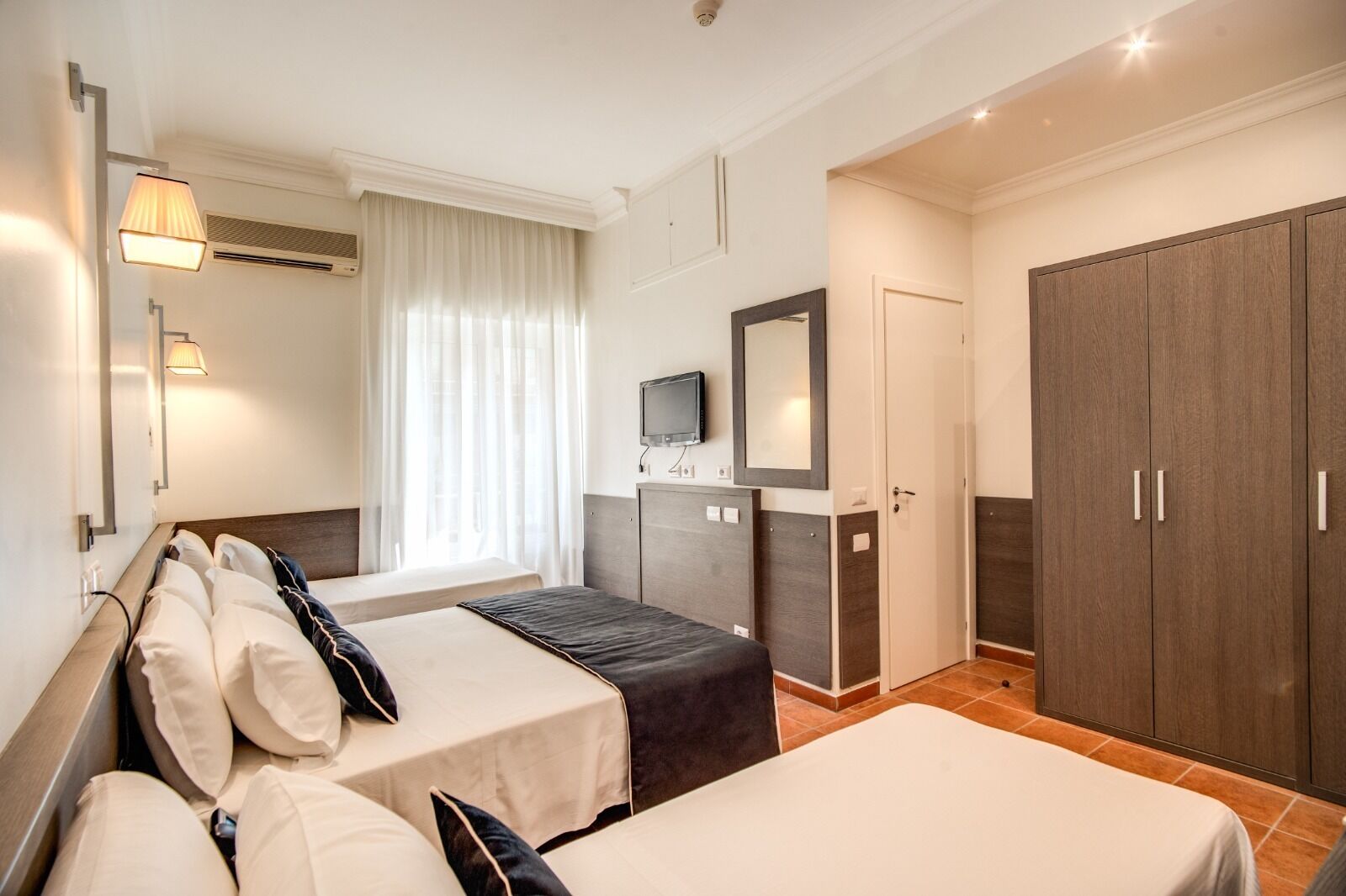 Quarto de hotel com duas camas, televisão, guarda-roupa e janela com cortinas.