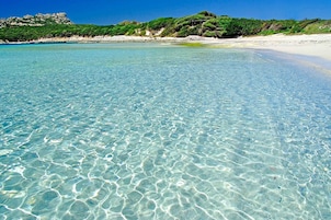 Mar azul-turquesa límpido com uma praia arenosa e afloramentos rochosos ao fundo.