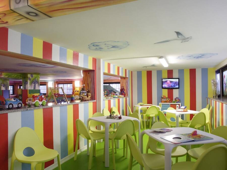 Espaço infantil com paredes coloridas listradas, cadeiras verdes e uma televisão fixada na parede.