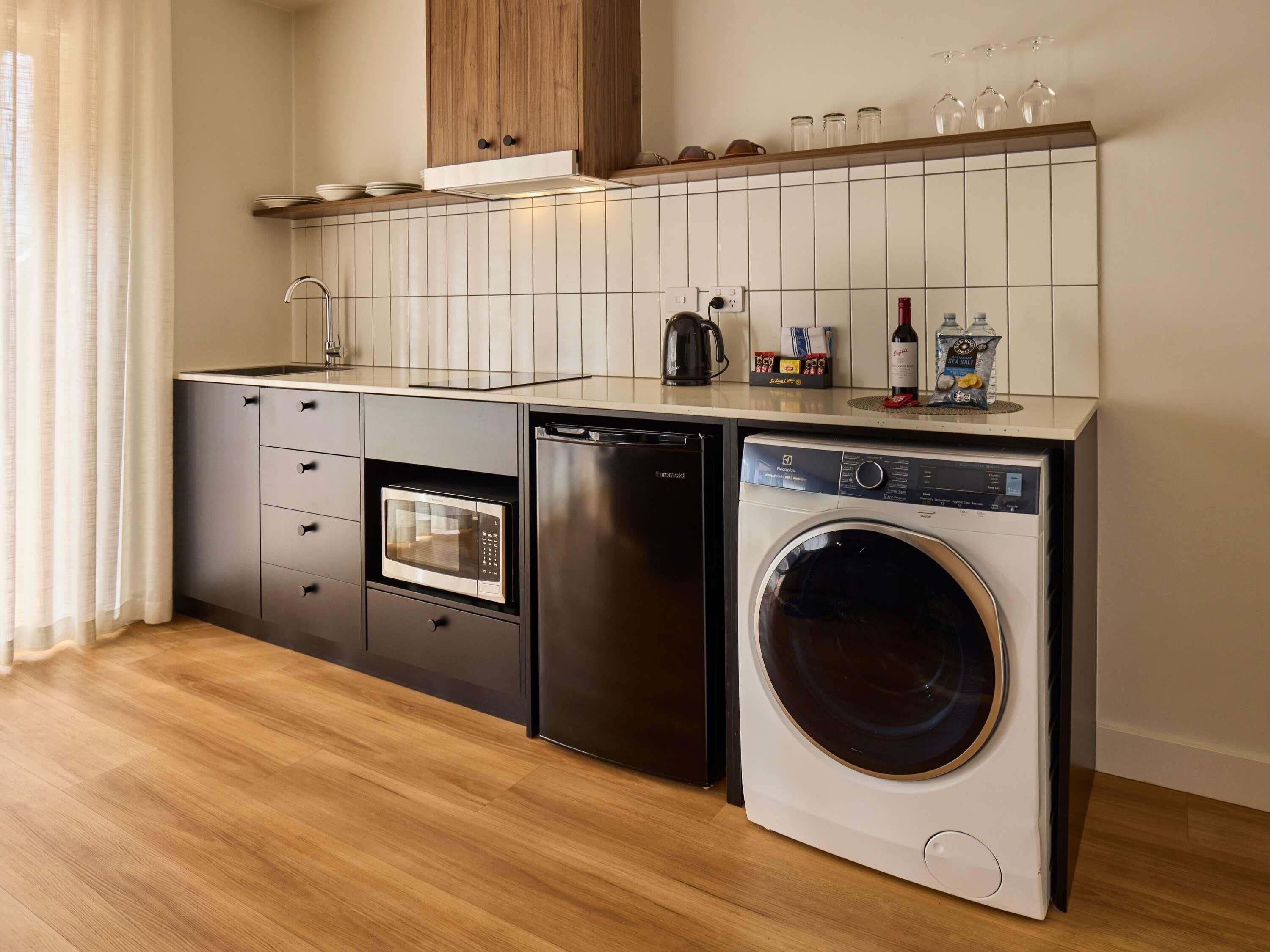 Cozinha compacta equipada com micro-ondas, lava-louças e máquina de lavar, armários de madeira e painel de azulejos.