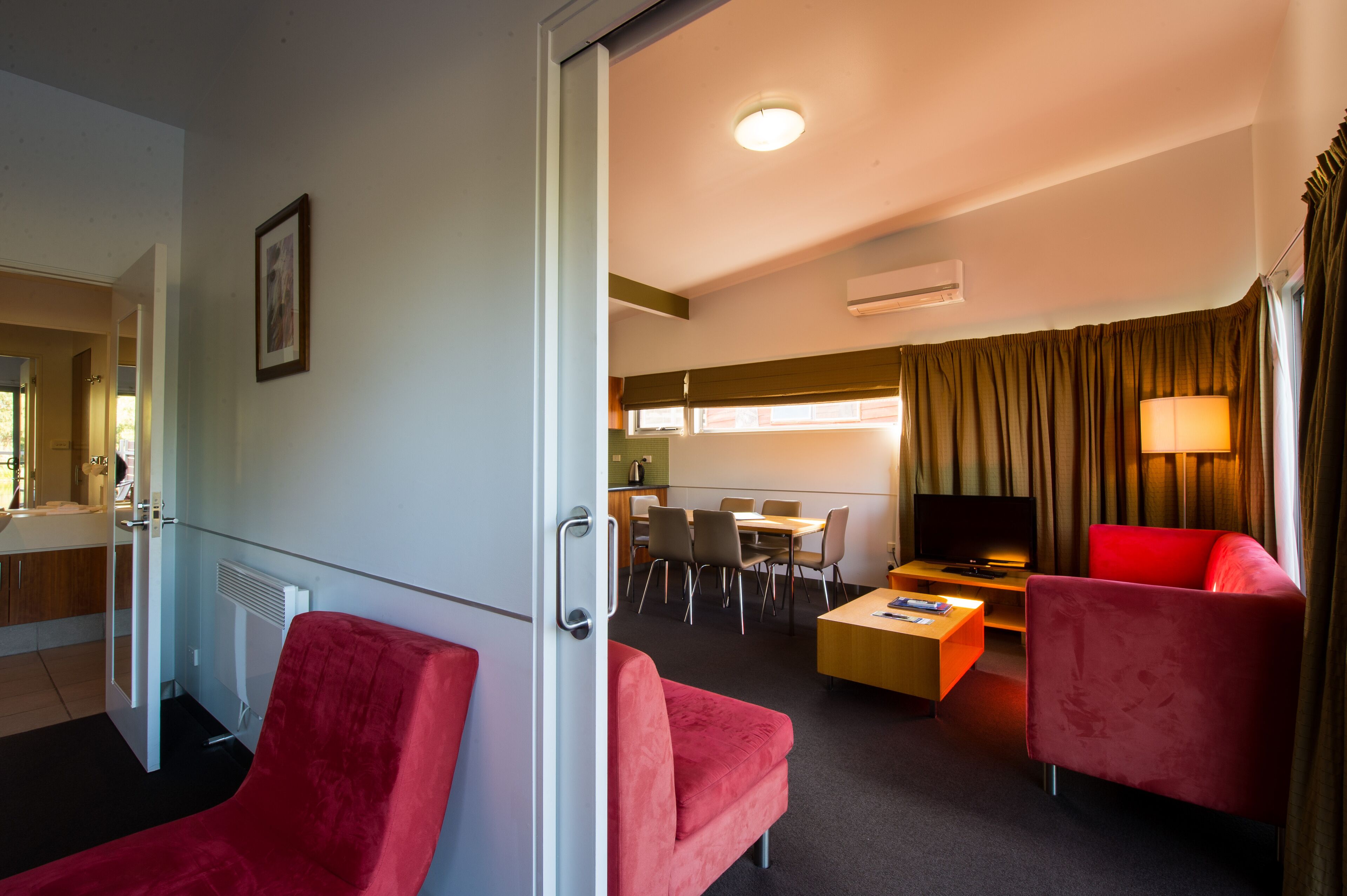 Quarto de hotel com sofá vermelho, poltrona vermelha, mesa de jantar com cadeiras, televisão e luminária.