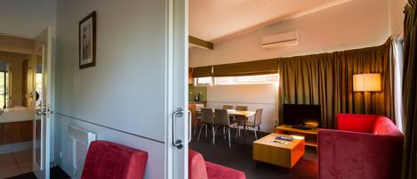 Quarto de hotel com sofá vermelho, poltrona vermelha, mesa de jantar com cadeiras, televisão e luminária.