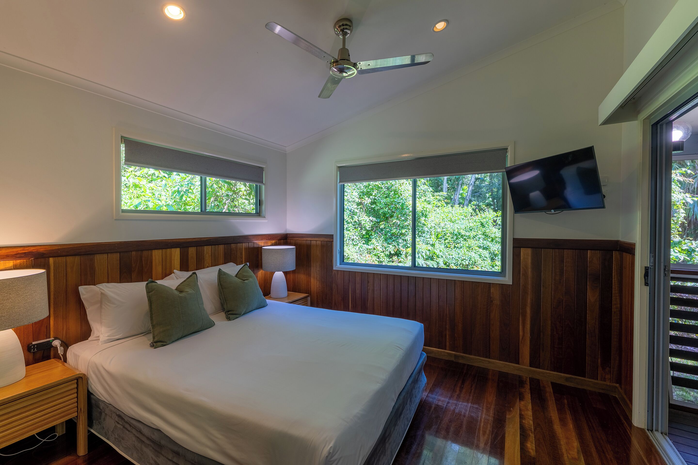 Quarto com cama, mesas de cabeceira, ventoinha no teto, duas janelas e televisão fixada na parede.