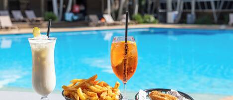 Mesa à beira da piscina com bebida, batatas fritas e salgados, com uma piscina e guarda-sóis ao fundo.