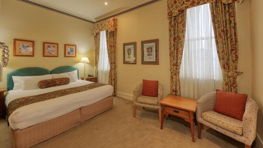 Quarto de hotel com cama, duas cadeiras, uma mesa e quadros nas paredes.