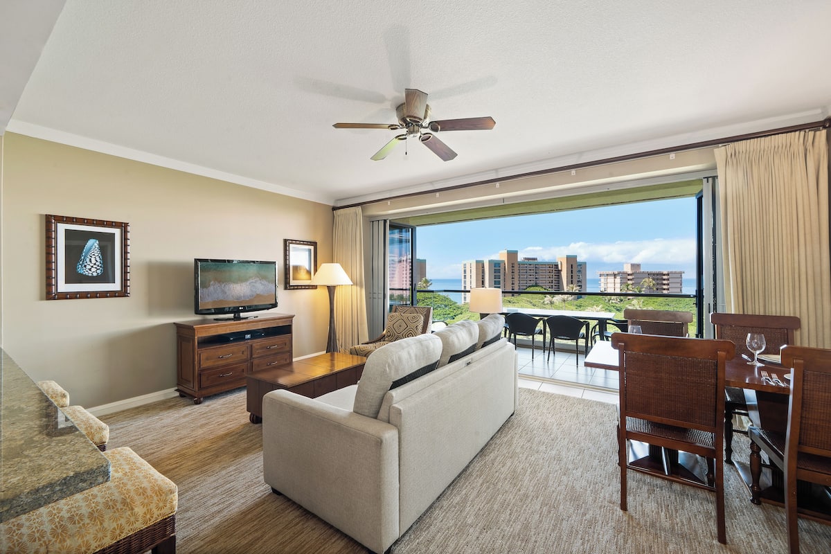Quarto de hotel com sofá, TV, mesa de jantar, cadeiras e varanda com vista para a cidade.