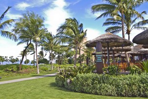 Resort tropical com construções de palha, palmeiras e um letreiro indicando café da manhã, almoço, jantar e coquetéis.
