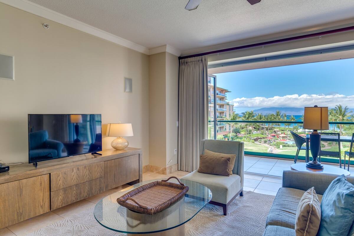 Quarto de hotel moderno com uma mesa de centro de vidro, televisão de tela plana, sofá e varanda com vista para o oceano e prédios.