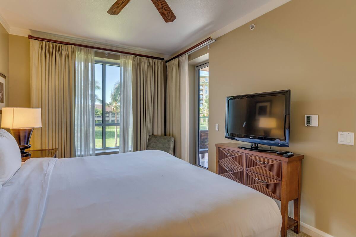 Quarto de hotel com uma cama grande, televisão sobre um suporte de madeira, uma cadeira e uma janela com cortinas.
