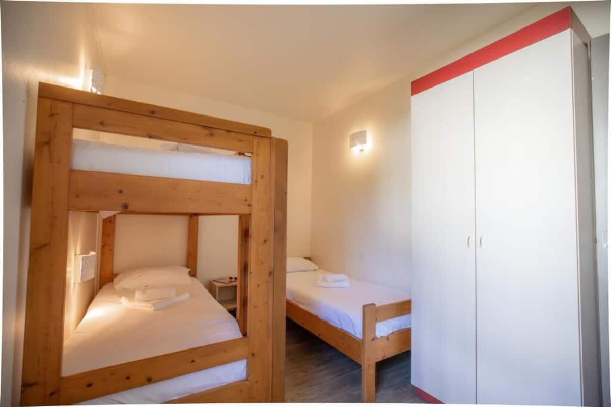 Quarto com duas beliches de madeira, um guarda-roupa branco e luminária fixada na parede.