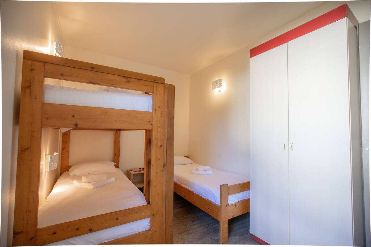 Quarto com duas camas beliches de madeira, um guarda-roupa branco e uma mesinha de cabeceira com uma lâmpada.