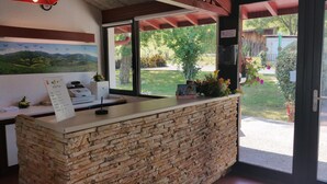 Balcão de recepção com fachada em pedra, área de recepção com computador e telefone, e vista para um jardim através de grandes janelas.