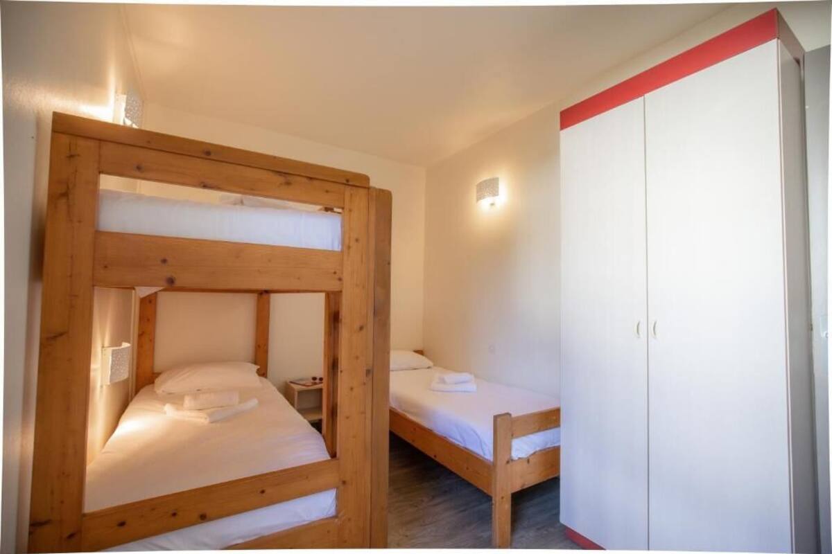 Quarto com duas beliches de madeira, um guarda-roupa branco e luminária fixada na parede.