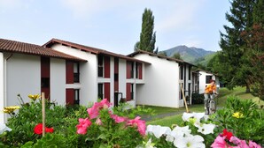 Fileira de edifícios brancos com persianas vermelhas, cercada por flores coloridas e vegetação.