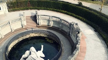 Uma fonte circular com uma estátua, rodeada por um caminho pavimentado e ladeada por sebes.