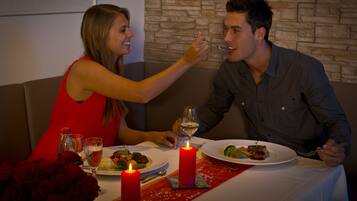 Um casal jantando à mesa, com velas, taças de vinho e um buquê de rosas.