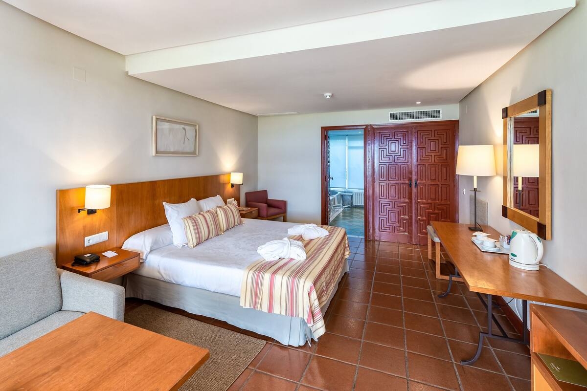 Quarto de hotel com cama, mesa de cabeceira, abajur, cadeira e escrivaninha com chaleira.