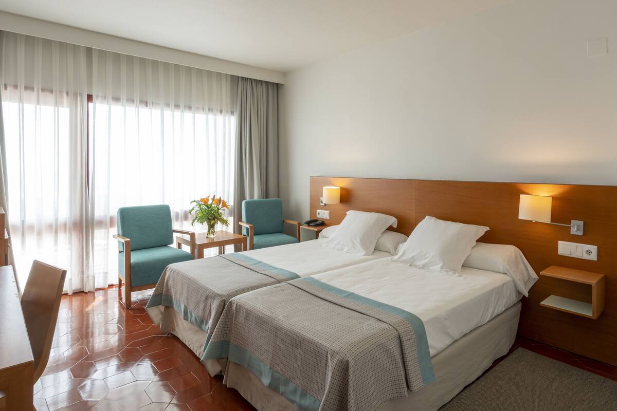 Quarto de hotel com duas camas, uma escrivaninha, uma cadeira, mesa de cabeceira e uma luminária.