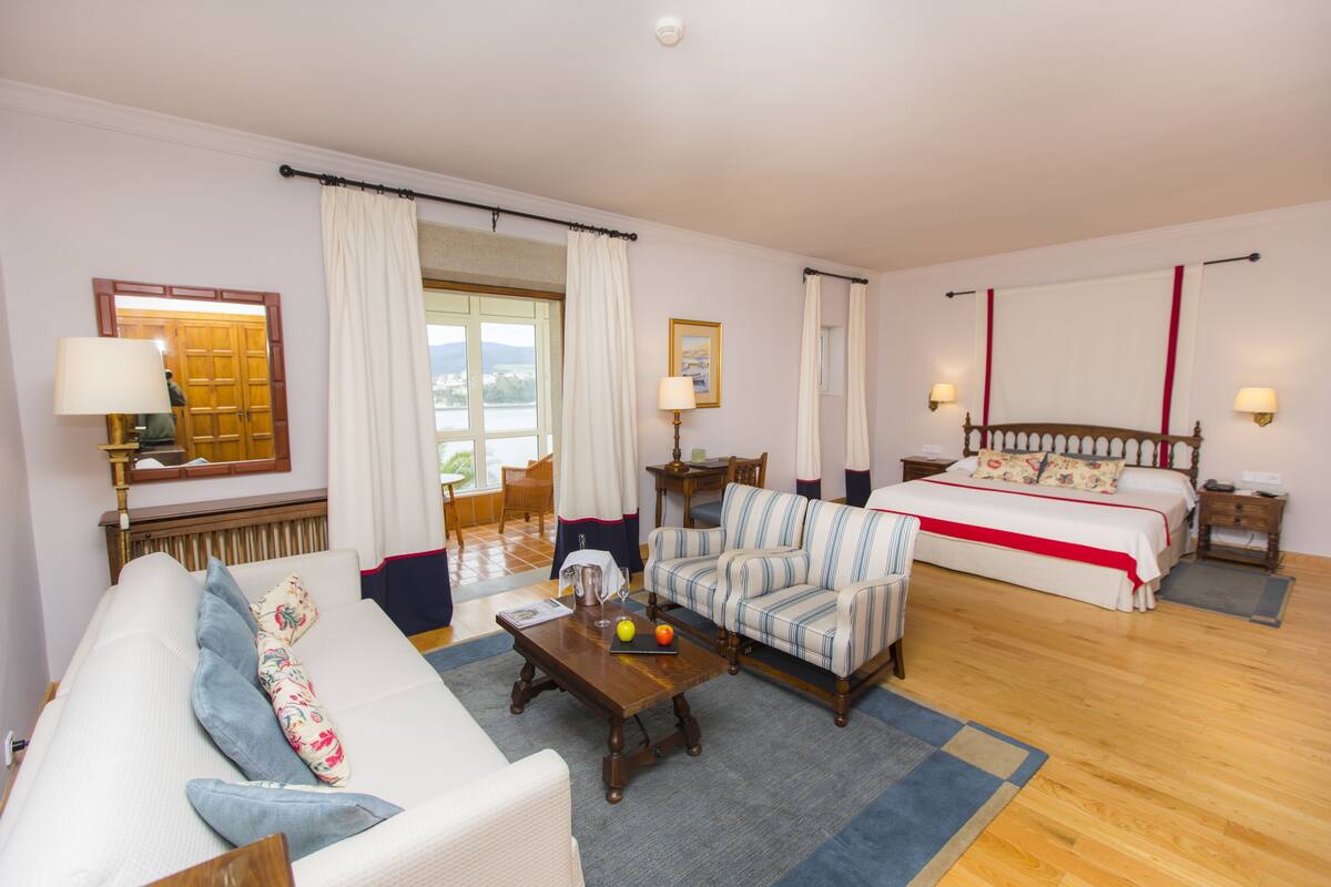 Quarto de hotel com cama, sofá, mesa de centro e janela com cortinas.