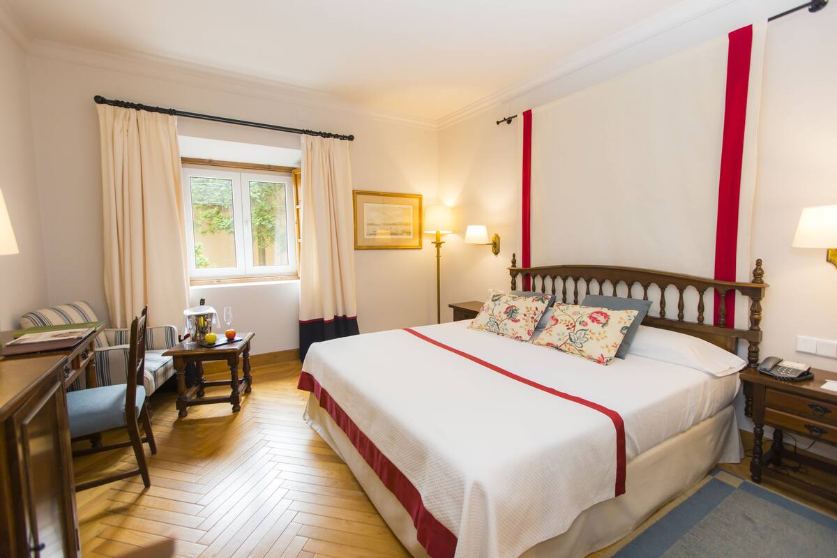 Quarto de hotel com uma cama grande, uma escrivaninha com cadeira, uma janela com cortinas e uma mesa de cabeceira com abajur.