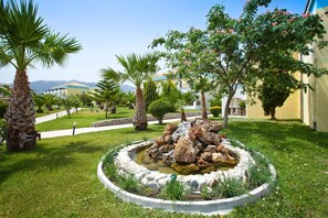 Um jardim ajardinado com um espelho d'água circular rodeado por pedras e plantas, palmeiras e um edifício com fachada amarela ao fundo.