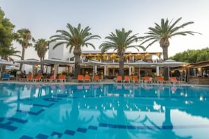 Piscina com espreguiçadeiras e palmeiras, cercada por um edifício com várias varandas.