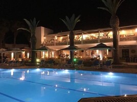 Piscina com espreguiçadeiras e guarda-sóis, um edifício com vários andares e área de estar ao ar livre, e palmeiras.