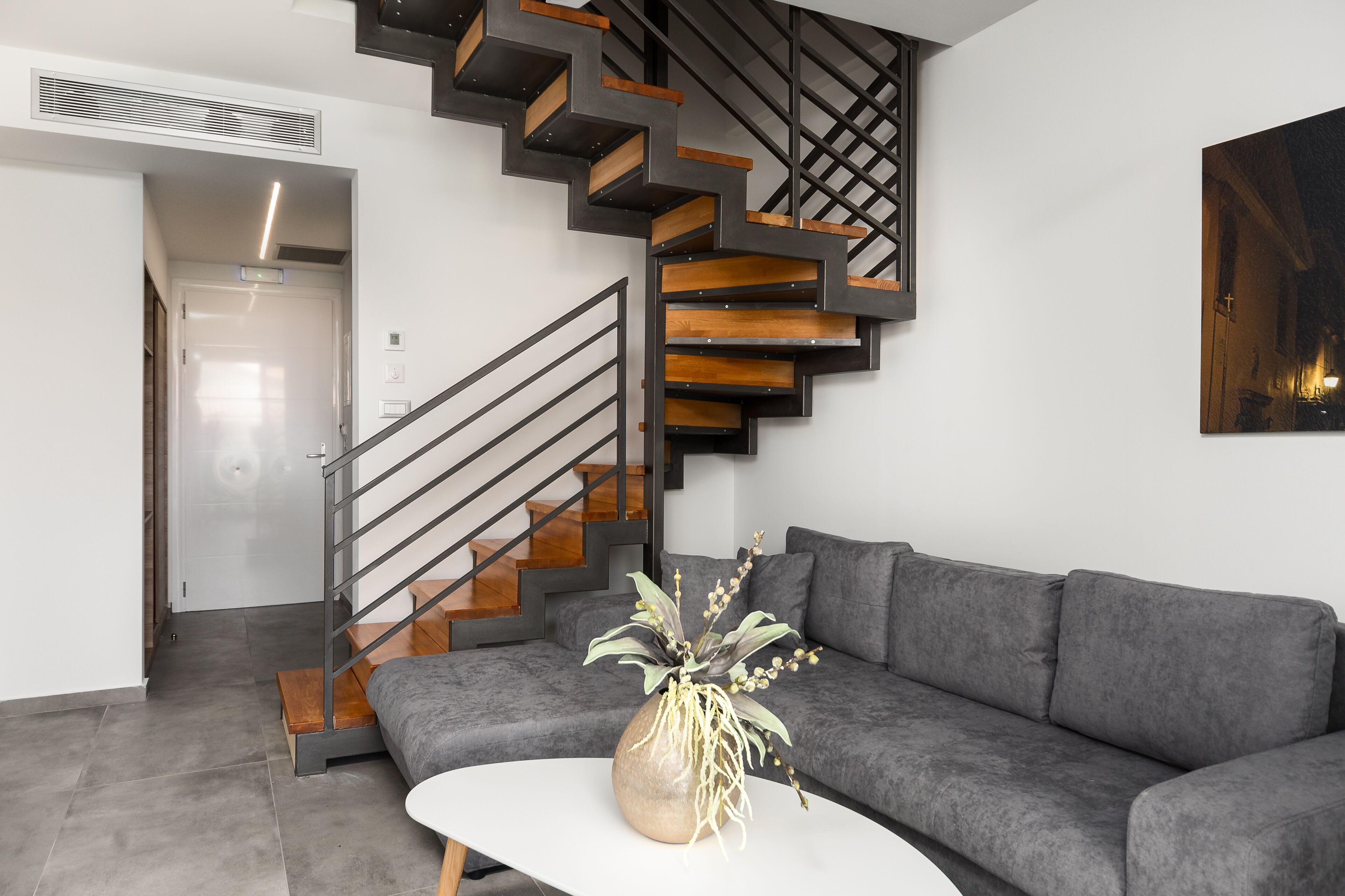 Uma sala de estar moderna com um sofá cinzento, uma mesa de centro branca e uma escada com degraus de madeira e corrimãos de metal.