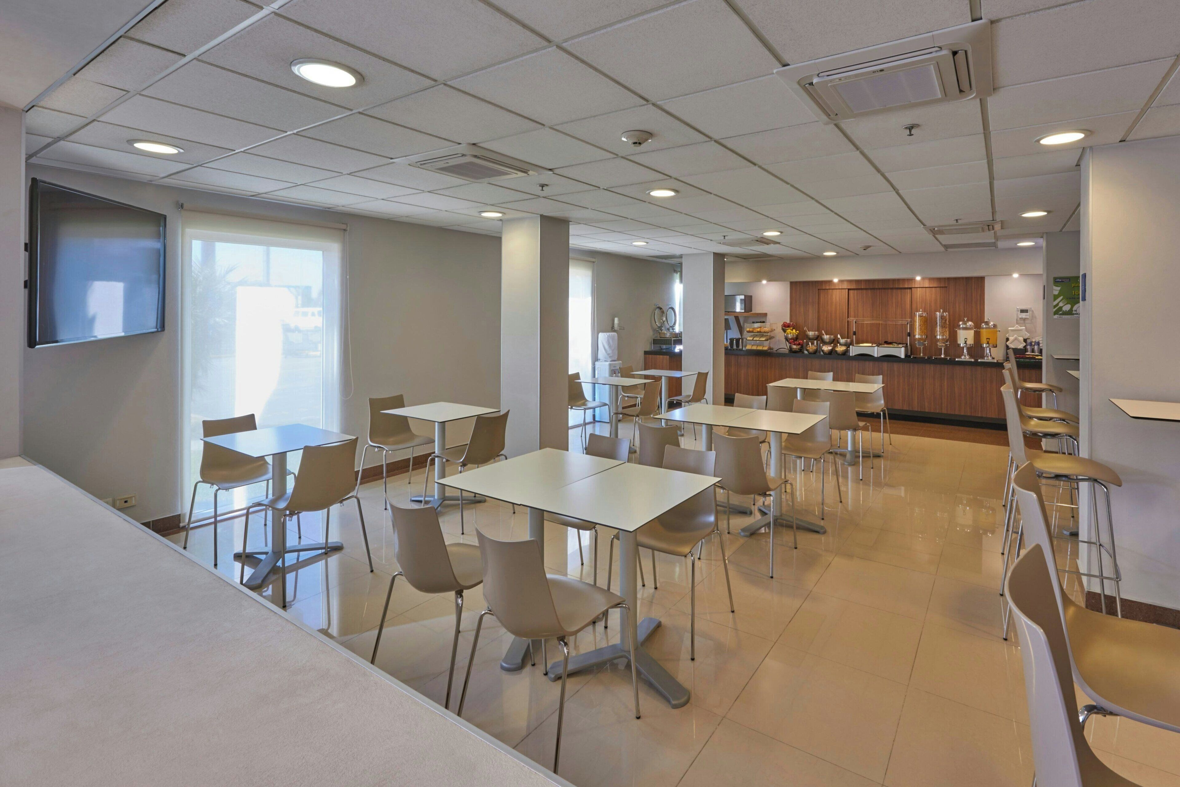 Uma sala de jantar moderna com mesas e cadeiras, uma cozinha ao fundo e uma grande janela que oferece iluminação natural.